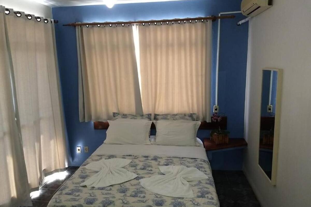 Quarto de hotel com cama, cortinas, mesa de cabeceira, espelho e ar condicionado fixado na parede.