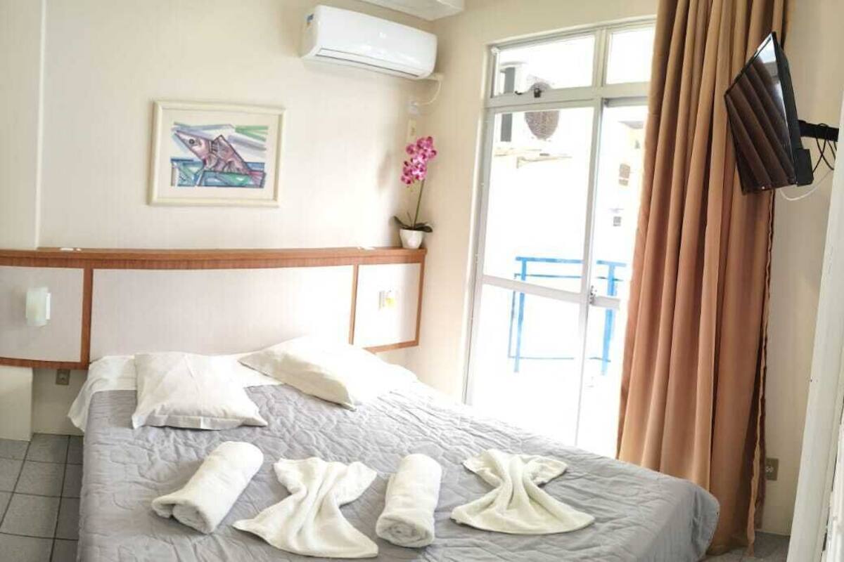 Quarto de hotel com cama, guarda-roupa de madeira, pequena mesa, ar condicionado na parede, quadro emoldurado e janela com cortinas.