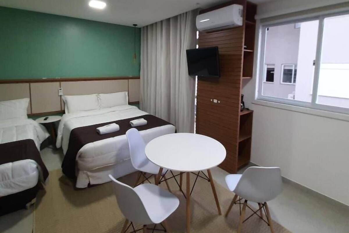 Quarto de hotel com cama, duas mesas de cabeceira, televisão, uma pequena mesa redonda com cadeiras e janela com cortinas.