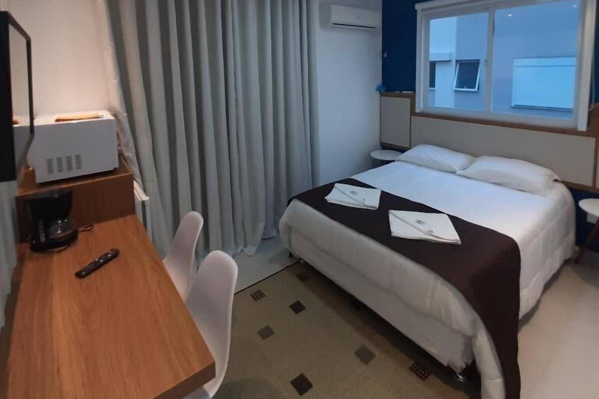 Quarto de hotel com uma cama, uma escrivaninha com computador, uma cadeira, uma janela e um ar condicionado.