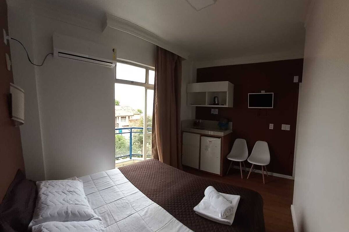 Quarto de hotel com uma cama, uma kitchenette compacta, duas cadeiras brancas e vista para o exterior.