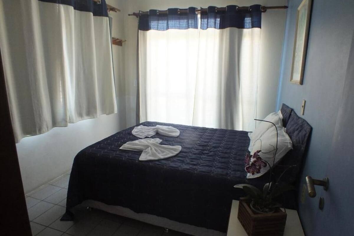 Quarto de hotel com cama, cortinas, mesa de cabeceira com uma planta e uma porta.