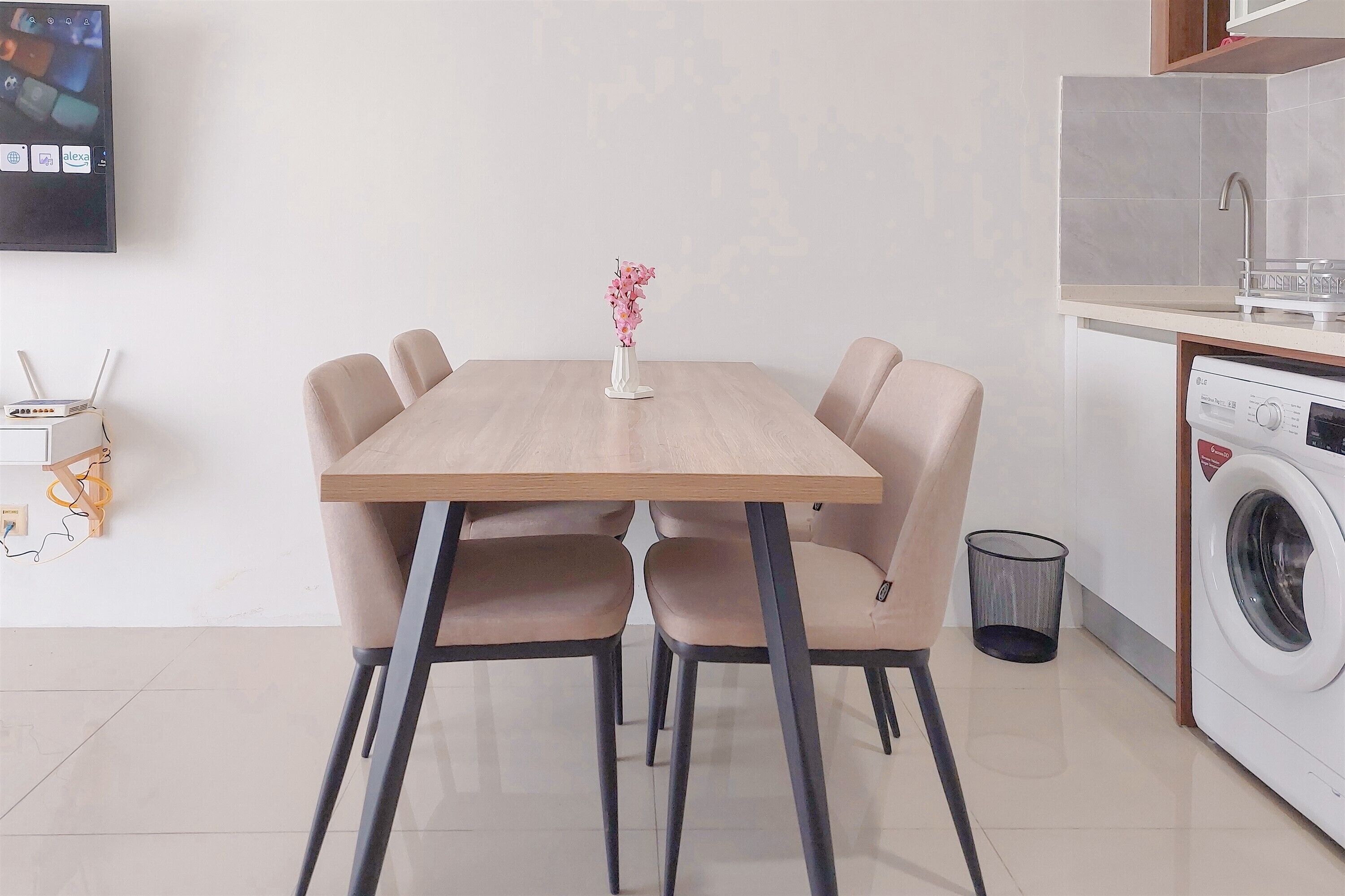 Uma área de refeições com mesa, cadeiras, um vaso com flores, uma máquina de lavar e uma TV na parede.