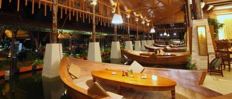 Um restaurante com mesas e cadeiras em formato de barco de madeira, teto alto com luminárias pendentes e ambiente acolhedor.