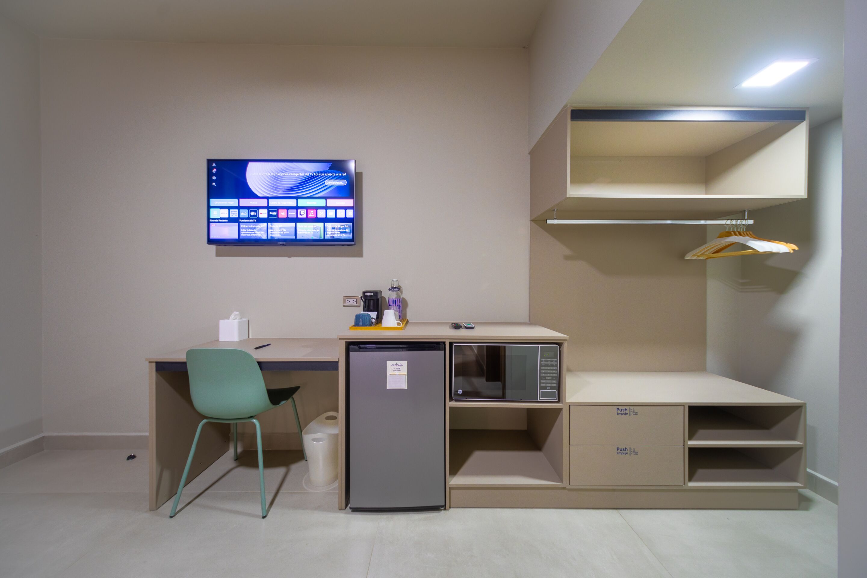 Quarto compacto com escrivaninha, cadeira, refrigerador, micro-ondas e televisão fixada na parede.