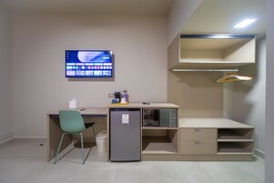 Quarto compacto com escrivaninha, cadeira, refrigerador, micro-ondas e televisão fixada na parede.