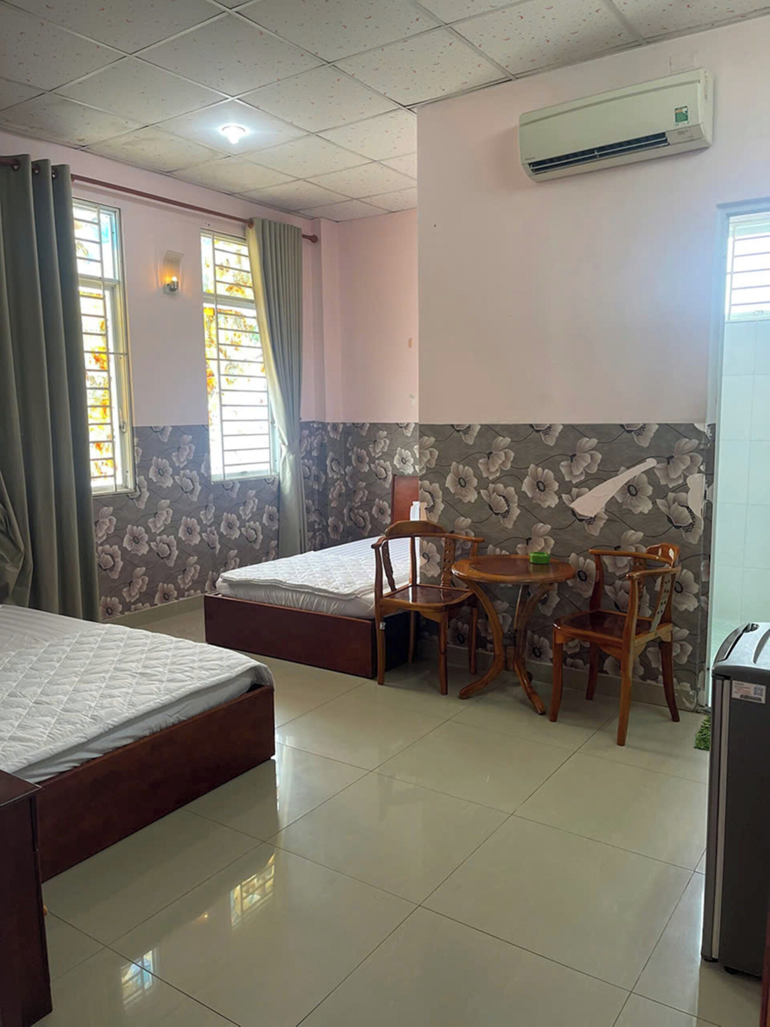 Quarto de hotel com duas camas, uma mesinha com cadeiras, ar condicionado e uma janela com cortinas.