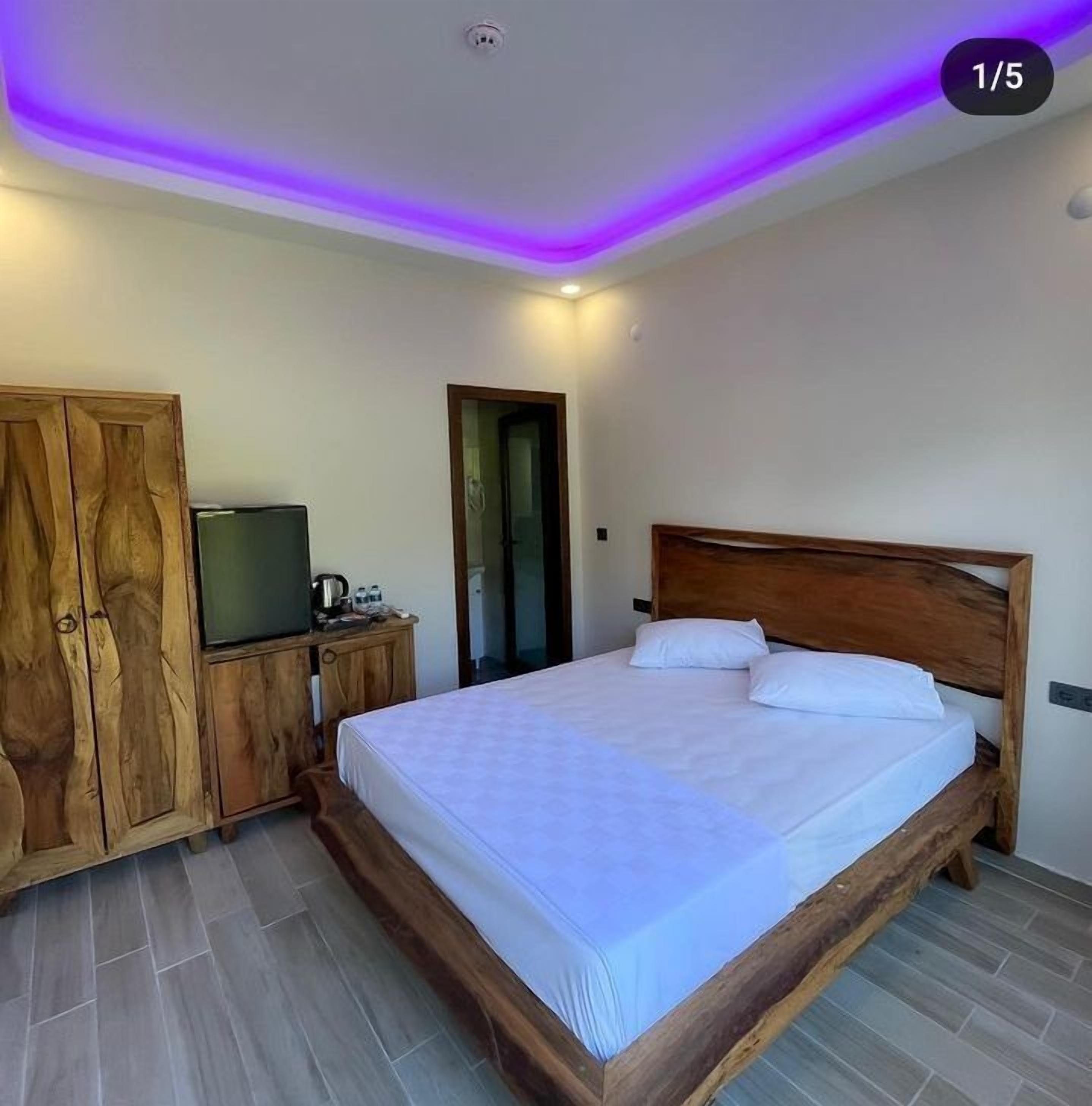 Quarto com estrutura de cama de madeira, TV em móvel de madeira e luminária de teto roxa.