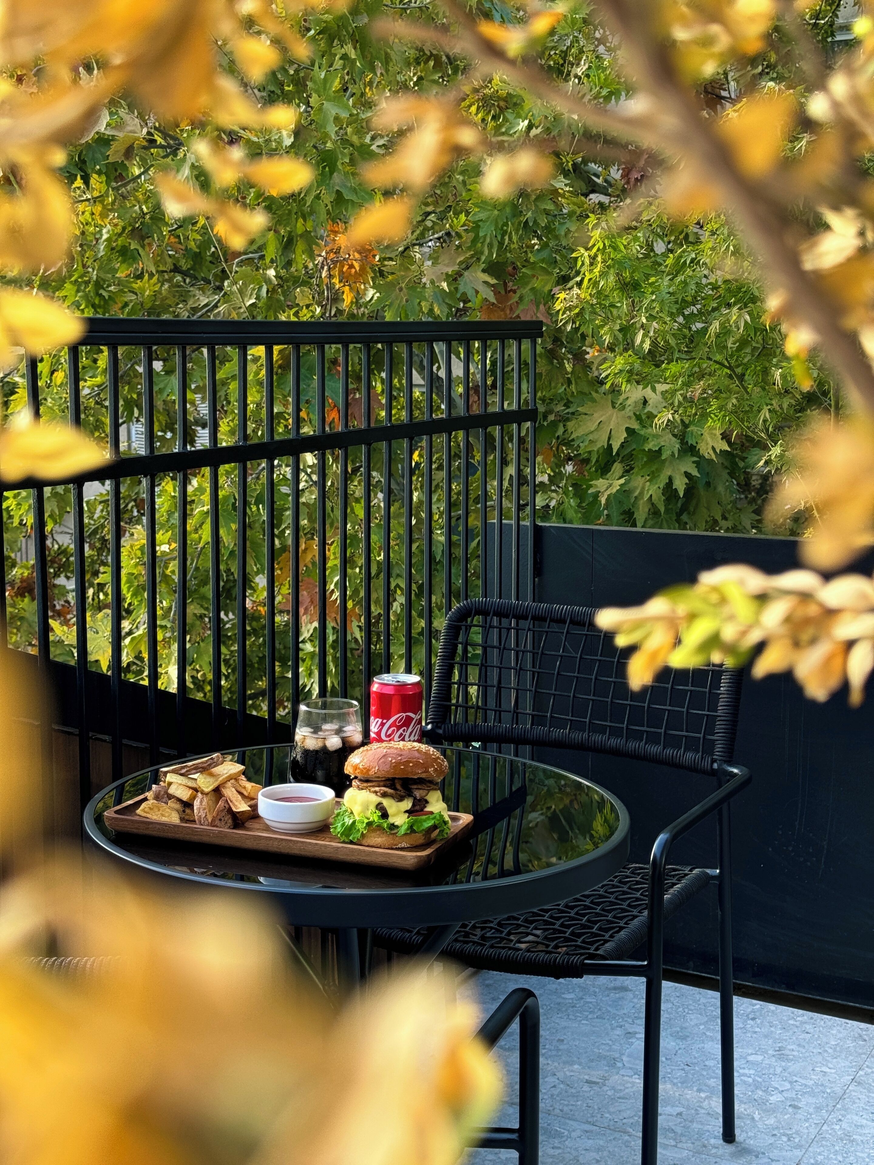 Uma mesa com comida e bebidas, uma cadeira, e vista para o verde através de uma cerca.