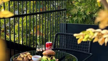 Uma mesa com comida e bebidas, uma cadeira, e vista para o verde através de uma cerca.