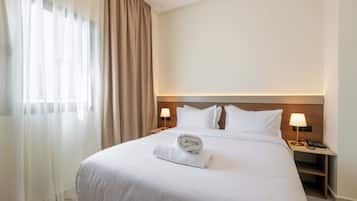 Quarto de hotel com cama bem arrumada, duas luminárias de cabeceira e janela com cortinas finas.