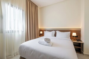 Quarto de hotel com cama bem arrumada, duas luminárias de cabeceira e janela com cortinas finas.