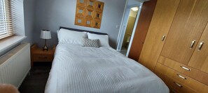 Quarto com cama, mesa de cabeceira, abajur, guarda-roupa de madeira e um enfeite na parede.