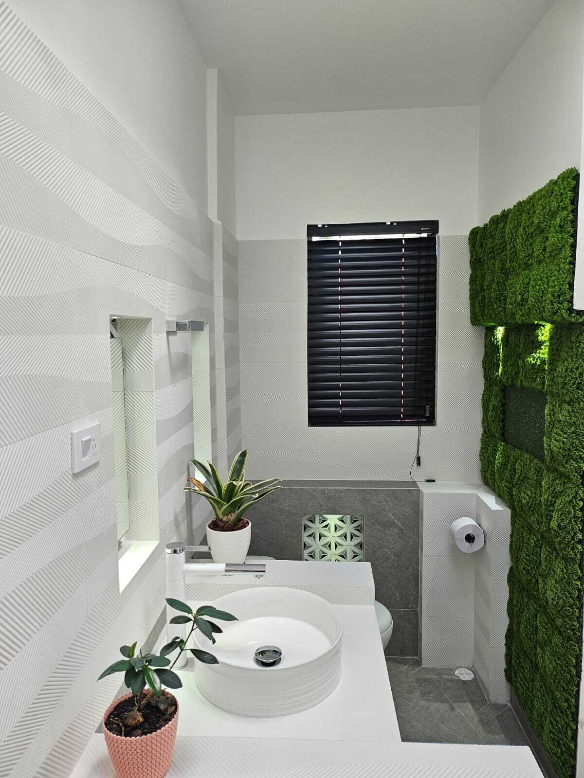Um banheiro moderno com pia branca, duas plantas em vasos, persiana preta e um painel verde na parede.
