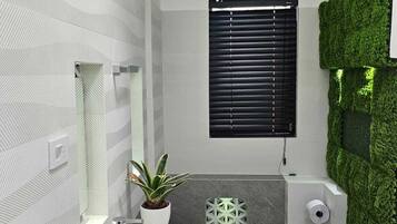 Um banheiro moderno com pia branca, duas plantas em vasos, persiana preta e um painel verde na parede.