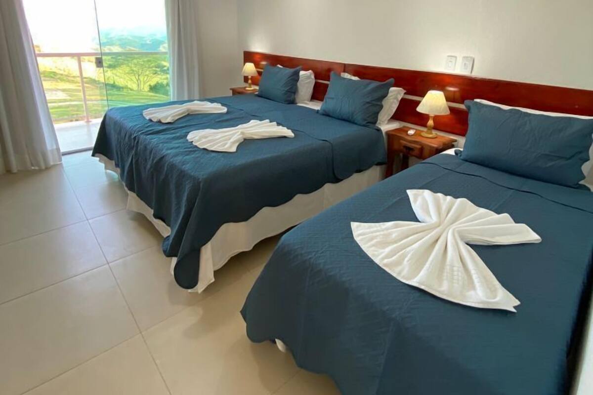 Quarto de hotel com duas camas, cada uma com colcha azul e toalhas brancas. Há um ventilador de teto, uma mesa de cabeceira com abajur e vista para o exterior através de uma grande janela.