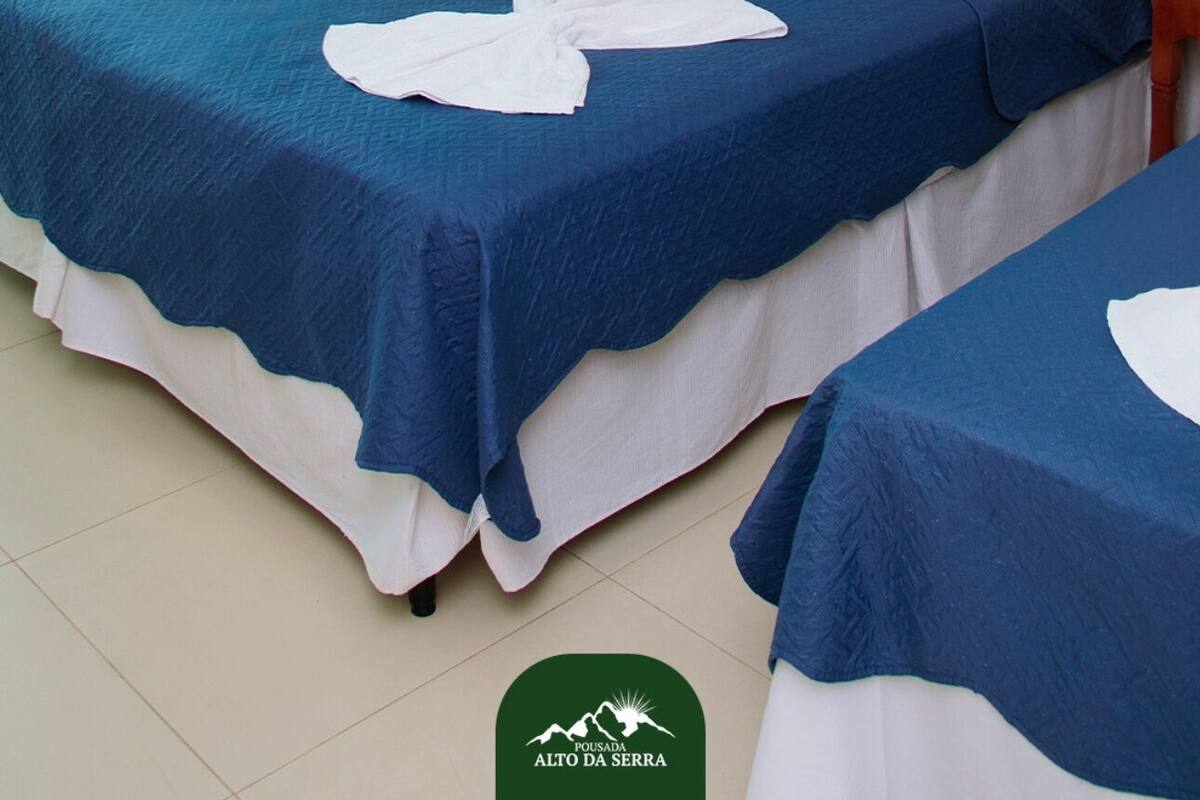 Cama de casal com roupa de cama azul e almofadas brancas, cabeceira de madeira e mesa de cabeceira com abajur.