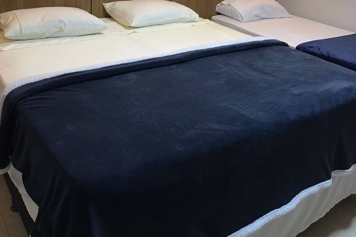 Quarto de hotel com duas camas, cada uma com roupa de cama branca e um cobertor azul escuro. O quarto possui uma cabeceira de madeira e uma luminária fixada na parede.