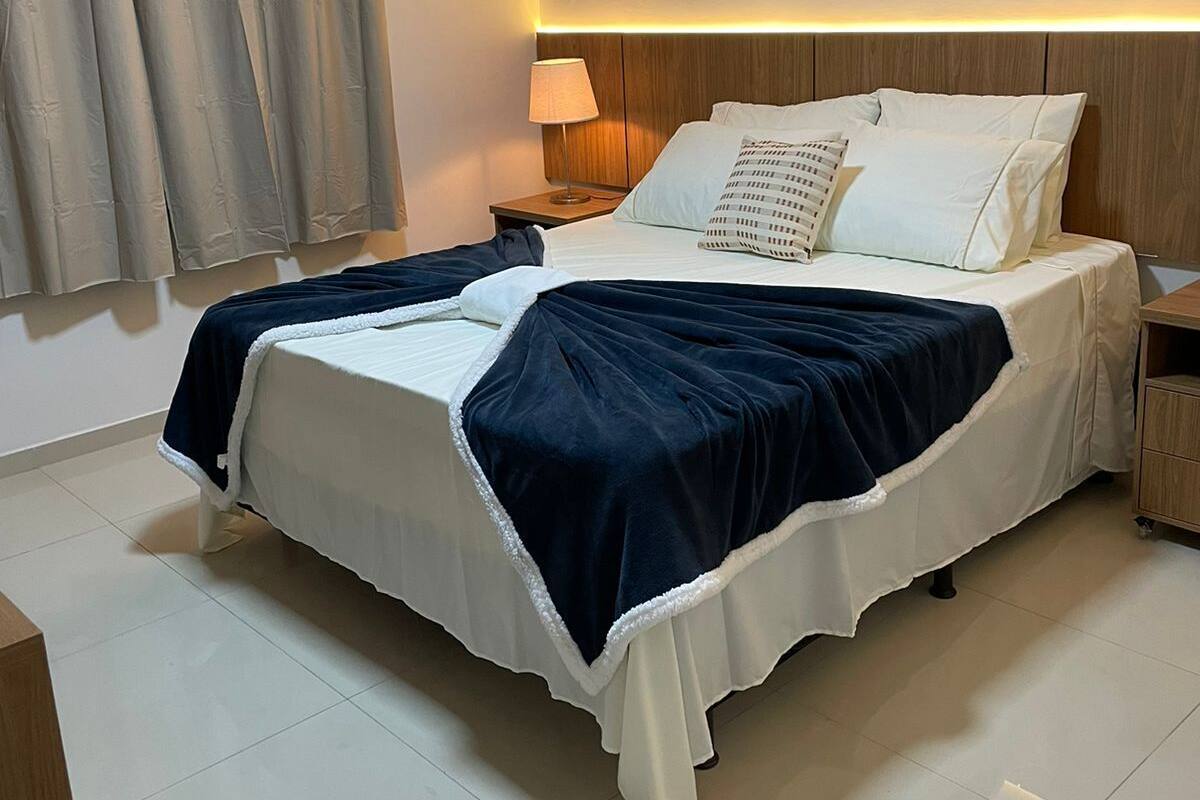 Quarto de hotel moderno com uma cama grande, mesa de cabeceira, abajur e vista para a cidade pela janela.
