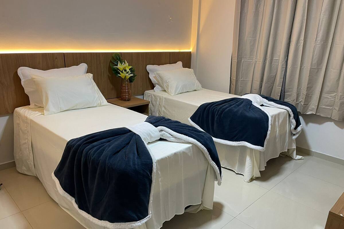 Quarto de hotel com duas camas, cada uma com roupa de cama branca e cobertores azul-marinho. Há uma pequena planta em uma mesinha de cabeceira, e o quarto possui uma cabeceira de madeira e uma cortina na janela.