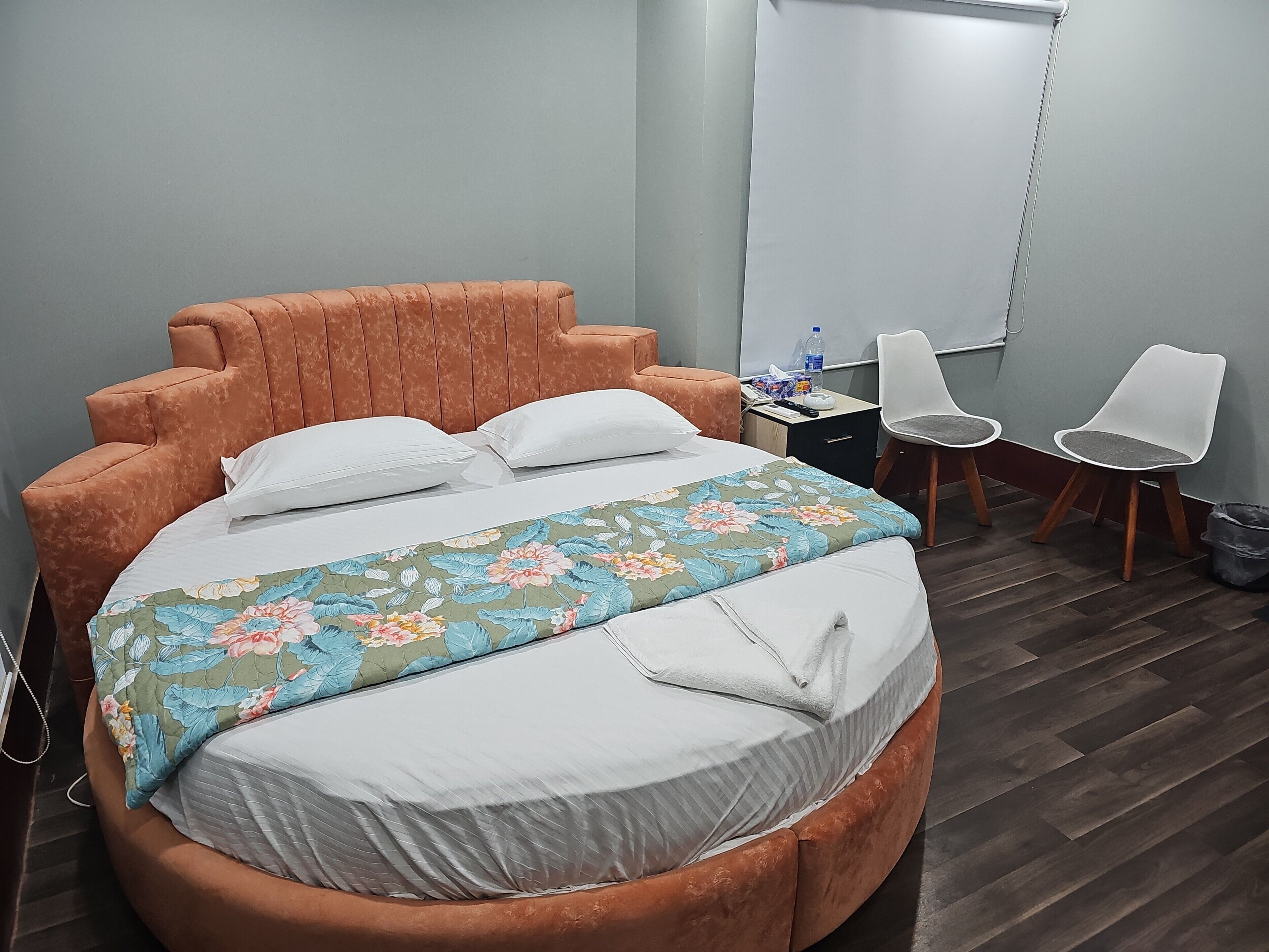Uma cama com colcha florida, duas almofadas brancas e cabeceira laranja. Há duas cadeiras brancas e uma escrivaninha com uma garrafa de água e um telefone.
