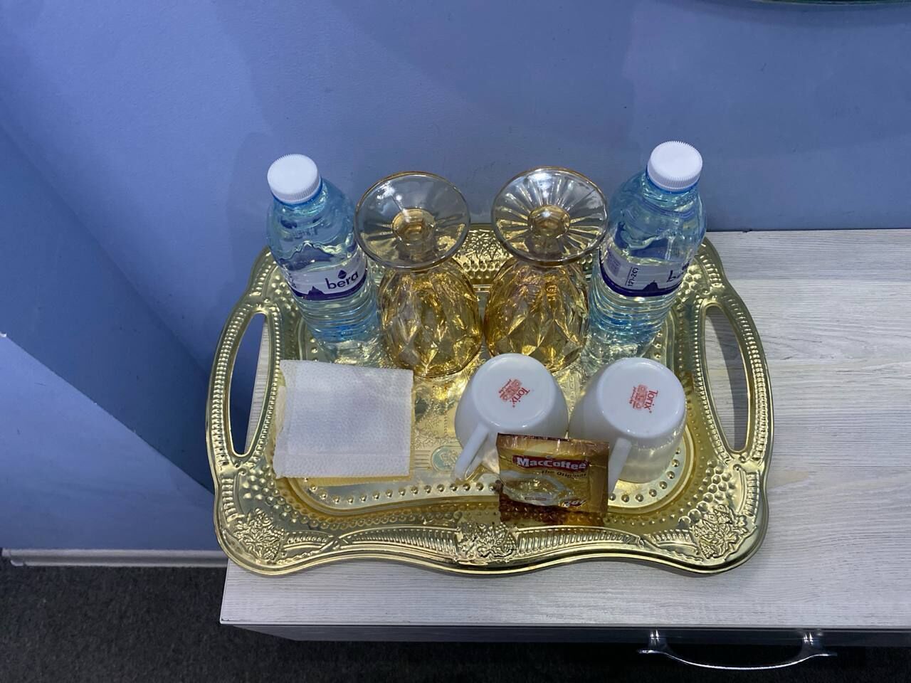 Uma bandeja com uma garrafa de água, uma garrafa de loção, uma garrafa de perfume, um lenço de papel e um sachê de MacCoffee.