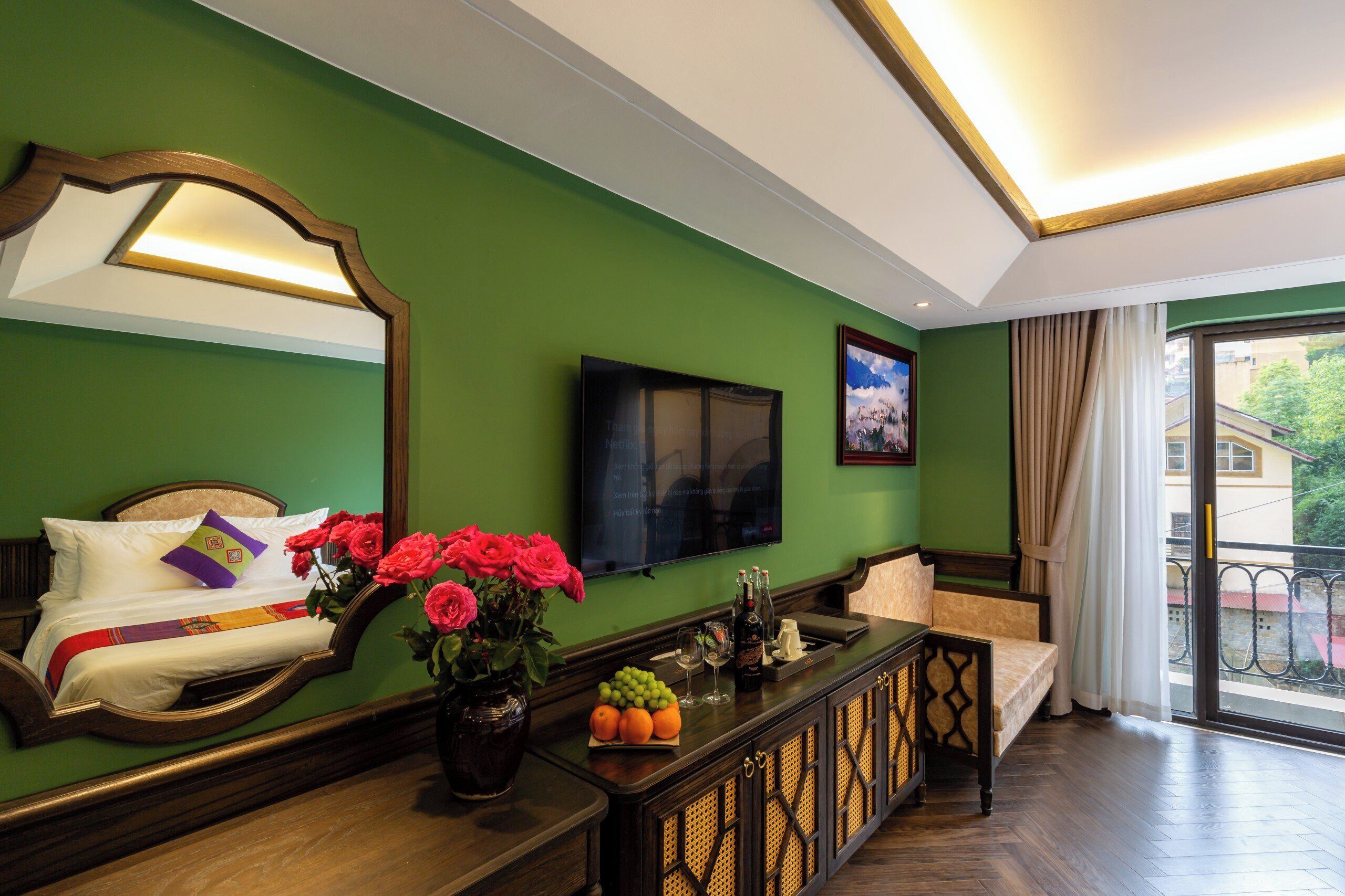 Quarto de hotel com parede verde, cama com cabeceira, um espelho grande, televisão de tela plana, uma mesa pequena com um vaso de flores e uma varanda com vista.
