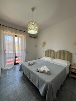 Cama bem arrumada com toalhas enroladas, luminária pendente e vista pela janela com cortinas leves.