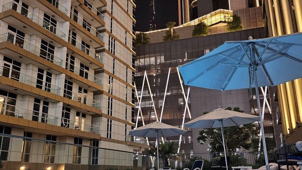 Uma piscina com espreguiçadeiras e guarda-sóis, rodeada por edifícios altos à noite.