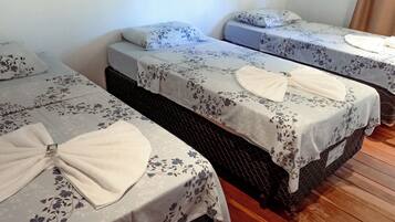 Quarto com três camas, cada uma com colcha estampada com flores e um laço na cama. O quarto possui piso de madeira e paredes brancas.