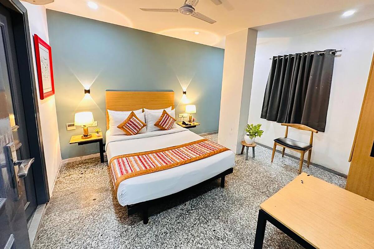 Quarto de hotel com cama, luminárias de cabeceira, ventilador de teto, mesinha, cadeira, planta em vaso e janela com cortinas.