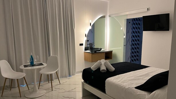 Quarto de hotel moderno com cama, duas cadeiras brancas, uma mesinha, espelho, televisão e banheiro com pia e espelho.