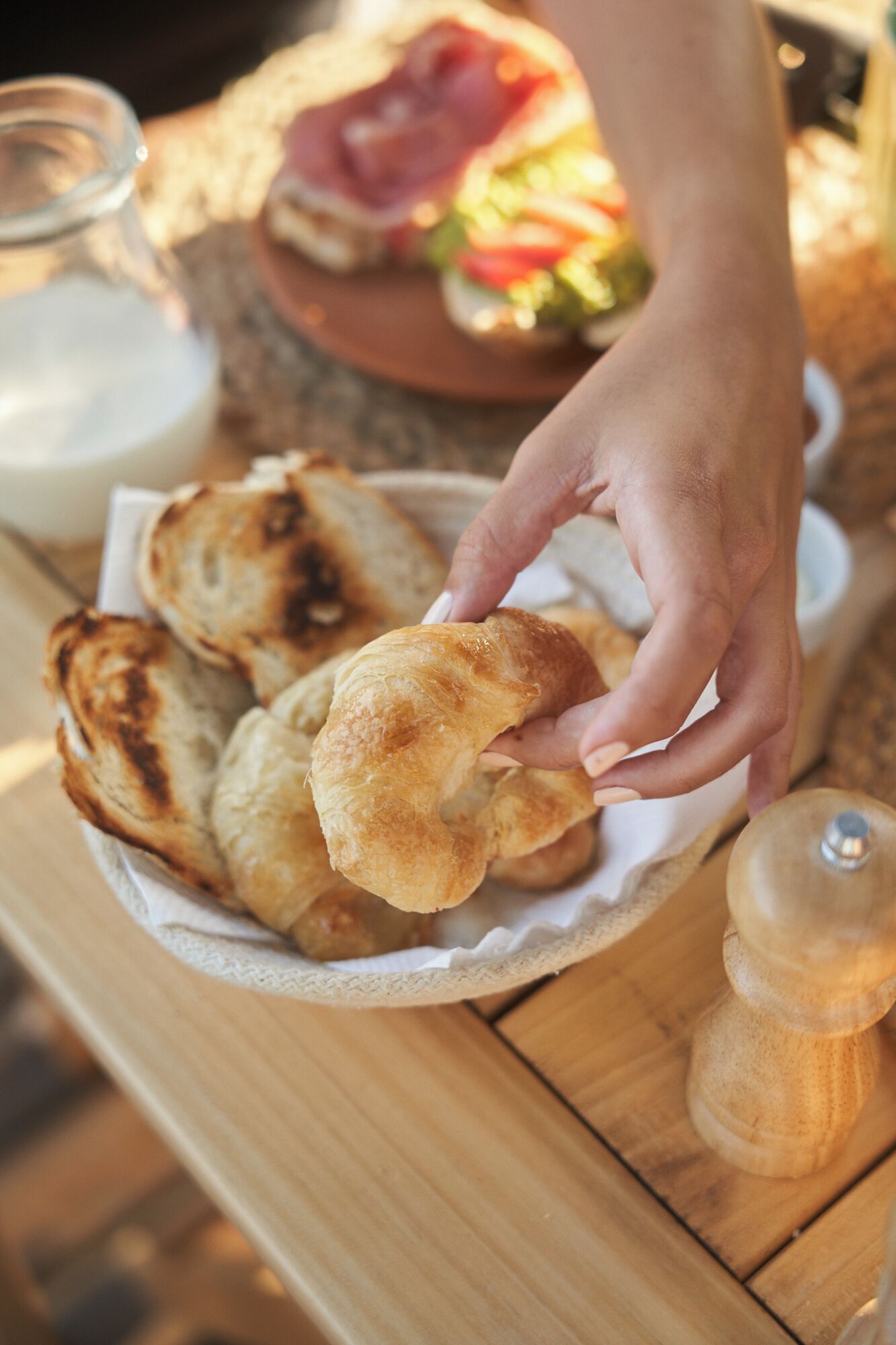 Mão segurando um croissant de uma cesta em uma mesa de madeira.