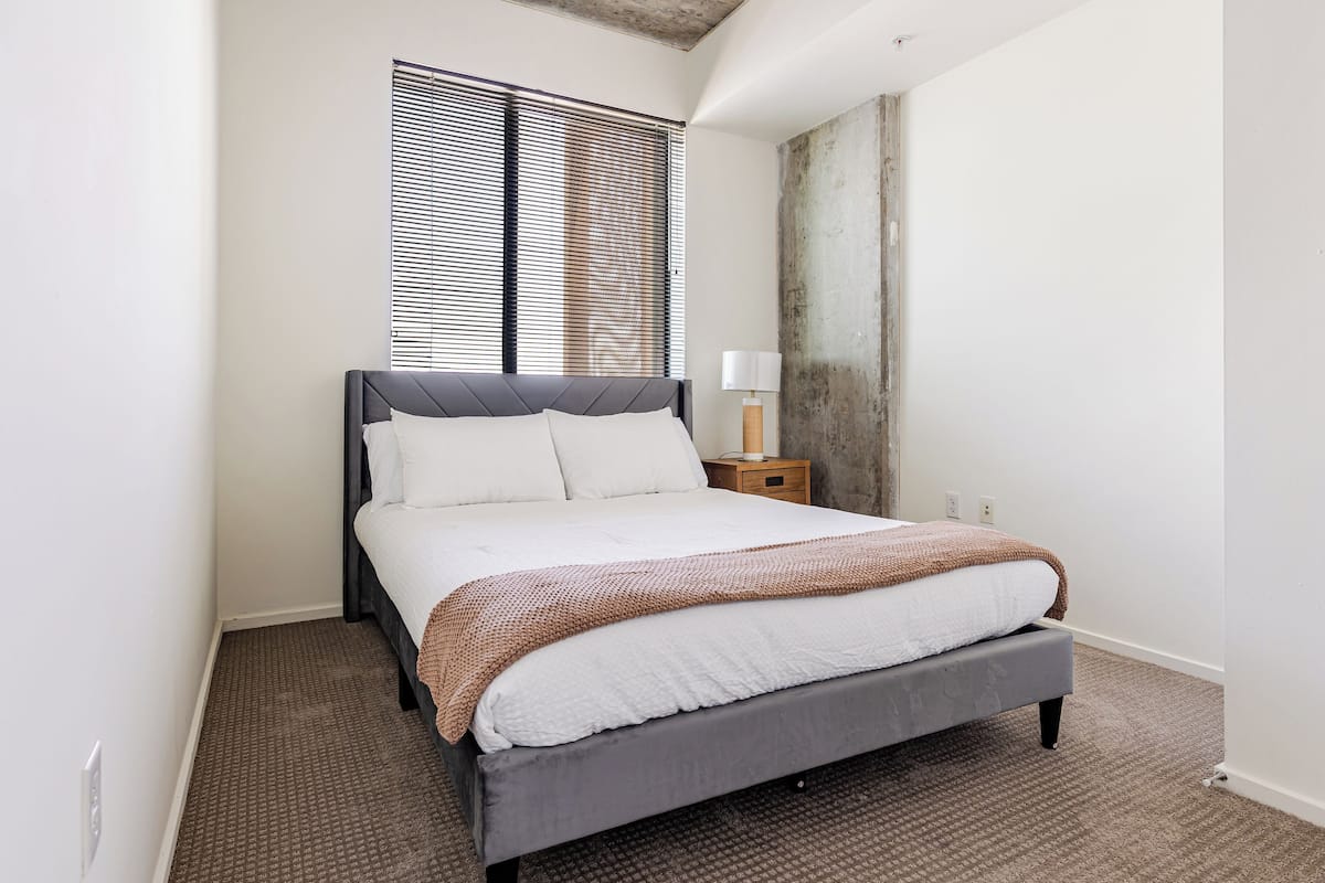 Uma cama bem arrumada com cabeceira cinza e roupa de cama branca, uma luminária de mesa e uma janela com persianas.