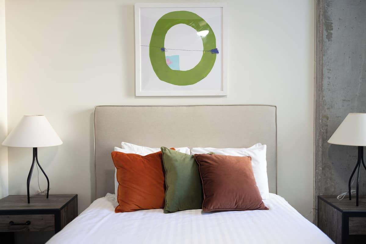 Cama bem arrumada com três almofadas, uma obra de arte emoldurada com uma letra 'O' verde grande na parede, e duas mesas de cabeceira com abajures.