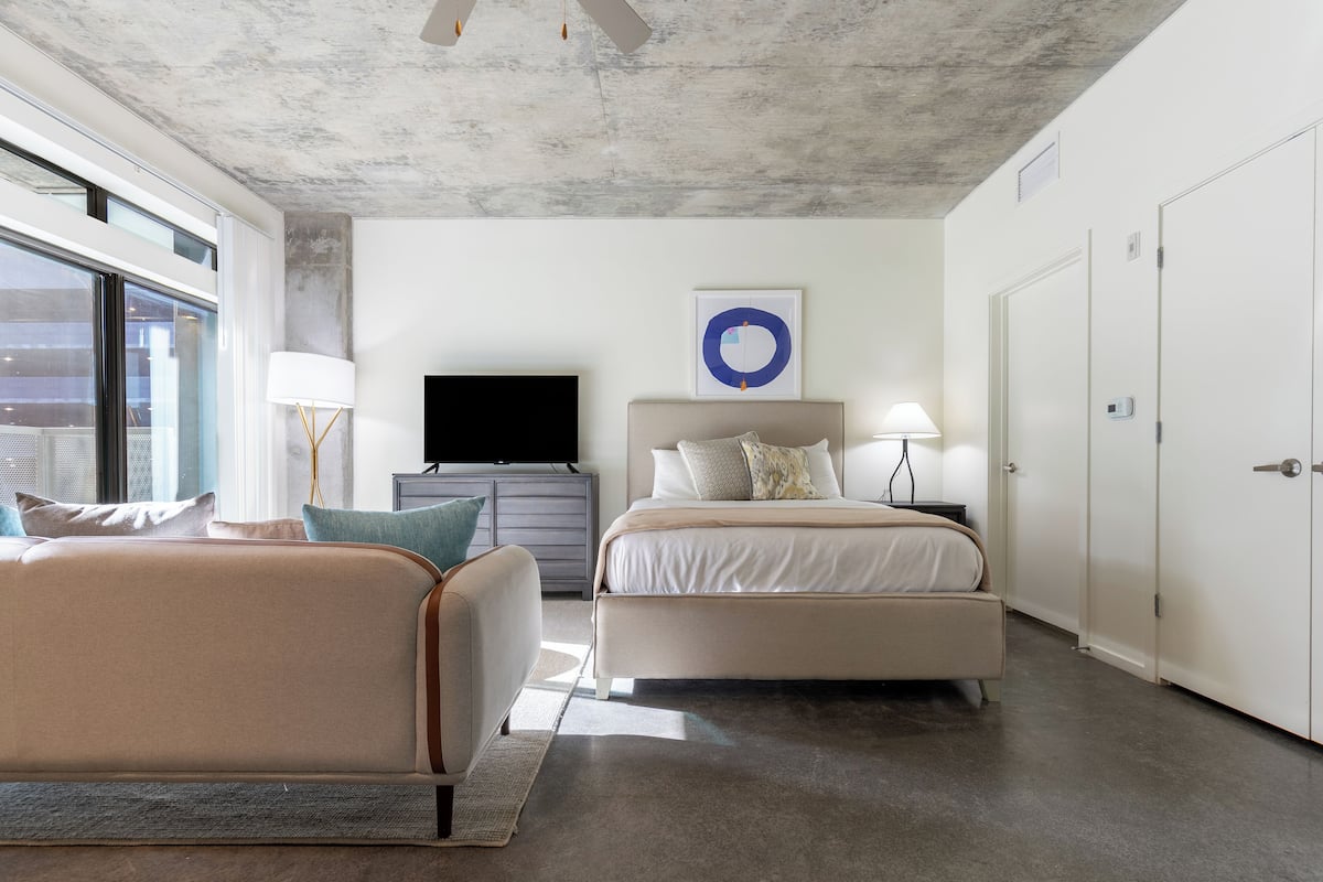 Quarto moderno com uma cama grande, um sofá menor, uma televisão de tela plana e uma janela ampla.