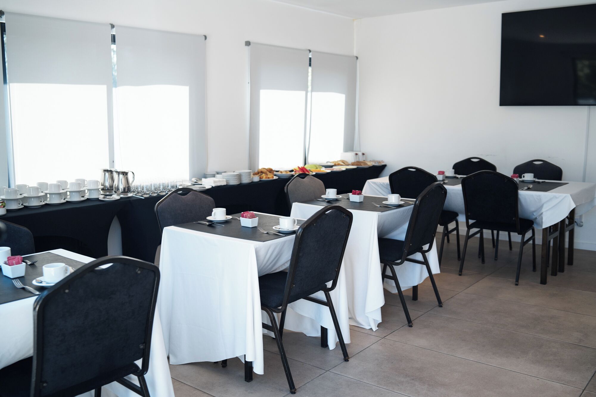 Uma área de refeições com mesas preparadas para o café da manhã, incluindo pratos, xícaras e talheres.