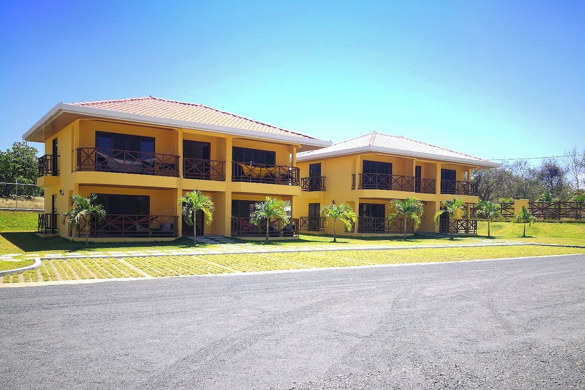 Um edifício amarelo de dois andares com varandas, calçada pavimentada e palmeiras.