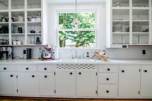 Uma cozinha com armários brancos, uma janela com vista para árvores e diversos eletrodomésticos.