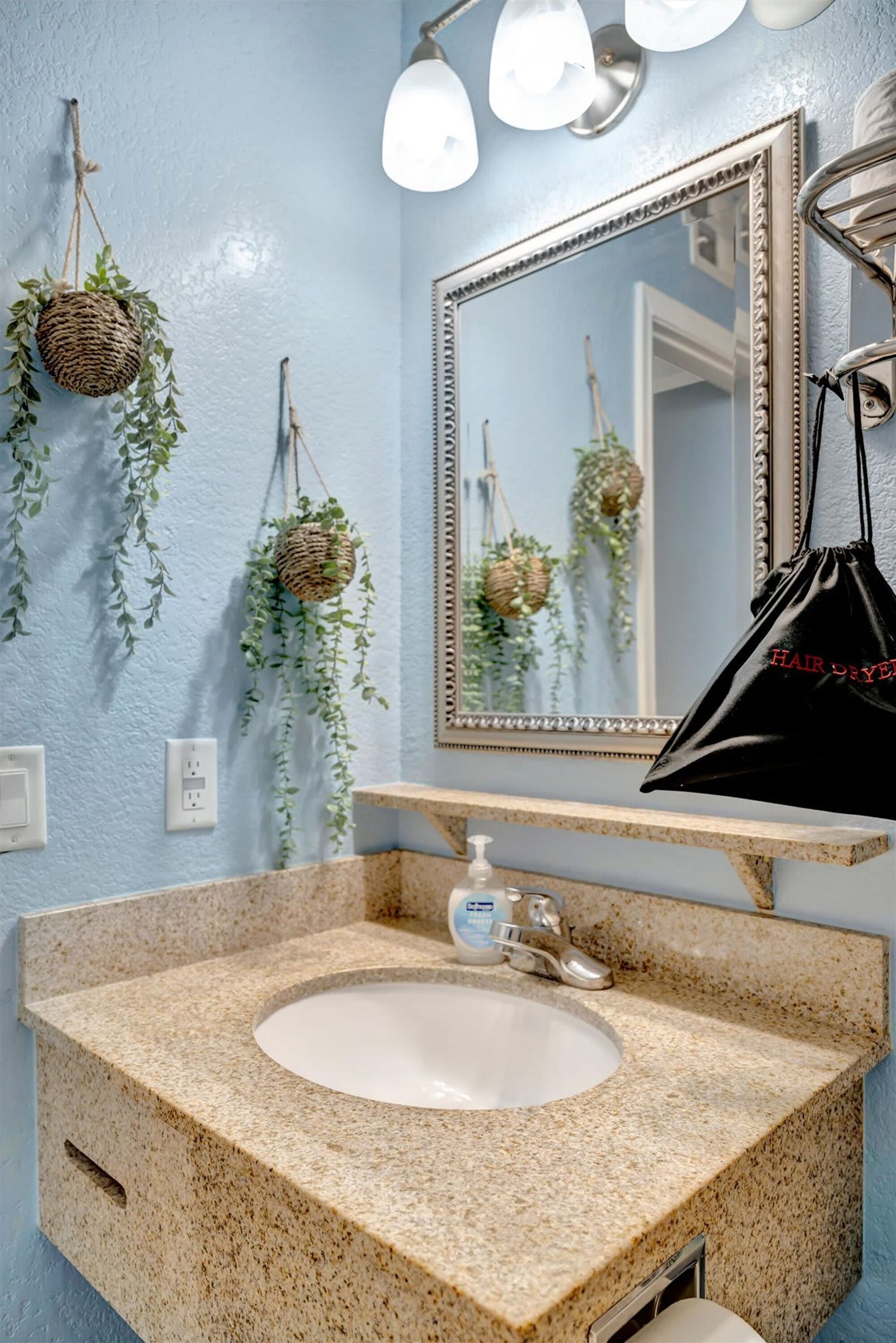 Casa de banho com lavatório de bancada em granito, espelho com moldura decorativa e plantas penduradas.