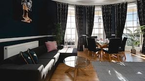 Uma sala de estar moderna com um sofá azul escuro, uma mesa de centro de vidro e uma área de jantar com mesa de madeira e cadeiras pretas.