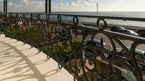 Varanda com grades de metal ornamentadas, vista para uma estrada costeira com carros estacionados e céu claro.