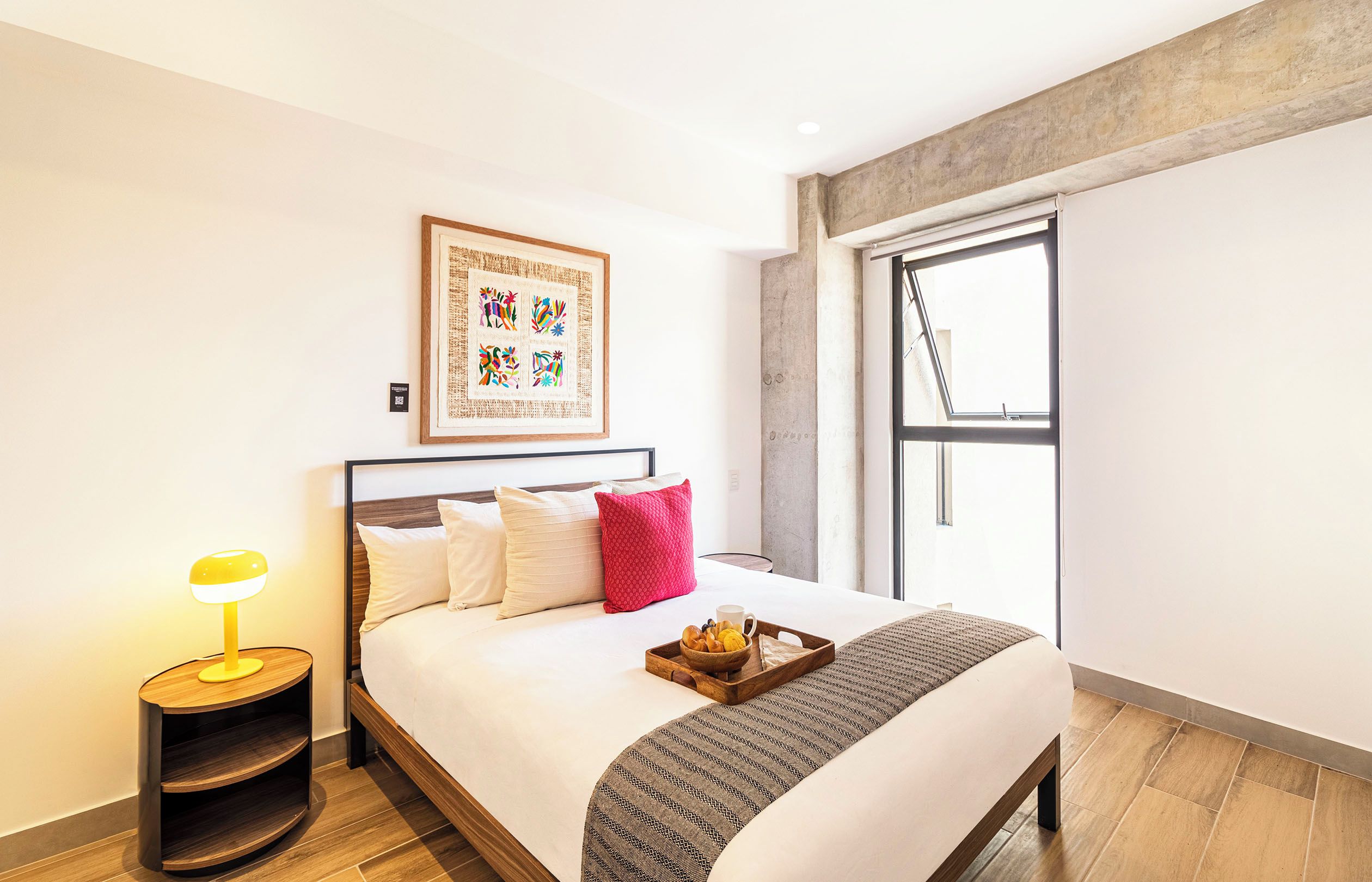 Quarto de hotel moderno com uma cama grande, mesa de cabeceira com abajur, uma obra de arte emoldurada acima da cama e janela com vista.