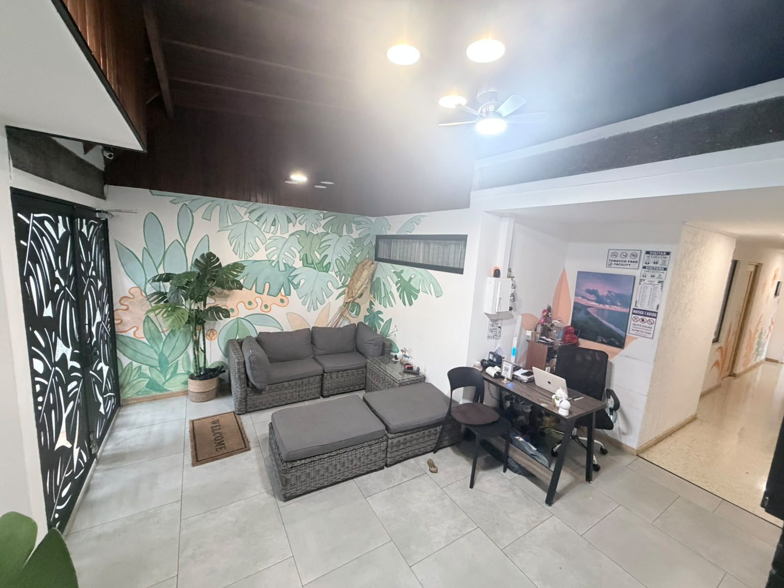 Espaço de estar com sofá, banqueta e escrivaninha com cadeira. Há uma planta grande e um mural na parede.