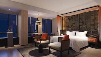Quarto de hotel moderno com uma cama grande, área de estar com uma mesa e vista para a cidade através de amplas janelas.