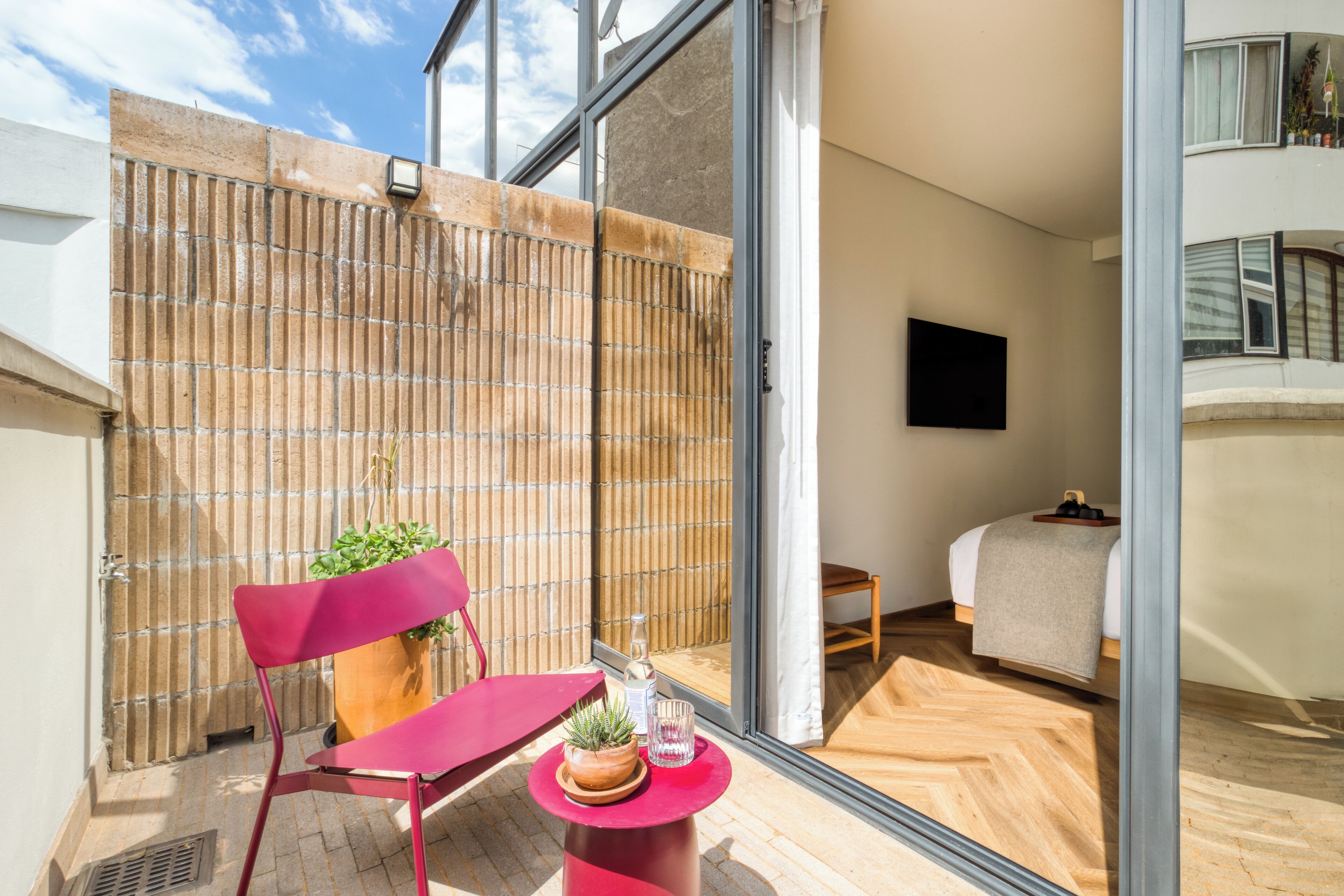 Um apartamento moderno com porta de vidro deslizante que dá para uma varanda, uma pequena área de estar com uma cadeira e mesa, e uma cama com mesa de cabeceira.