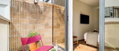 Um apartamento moderno com porta de vidro deslizante que dá para uma varanda, uma pequena área de estar com uma cadeira e mesa, e uma cama com mesa de cabeceira.