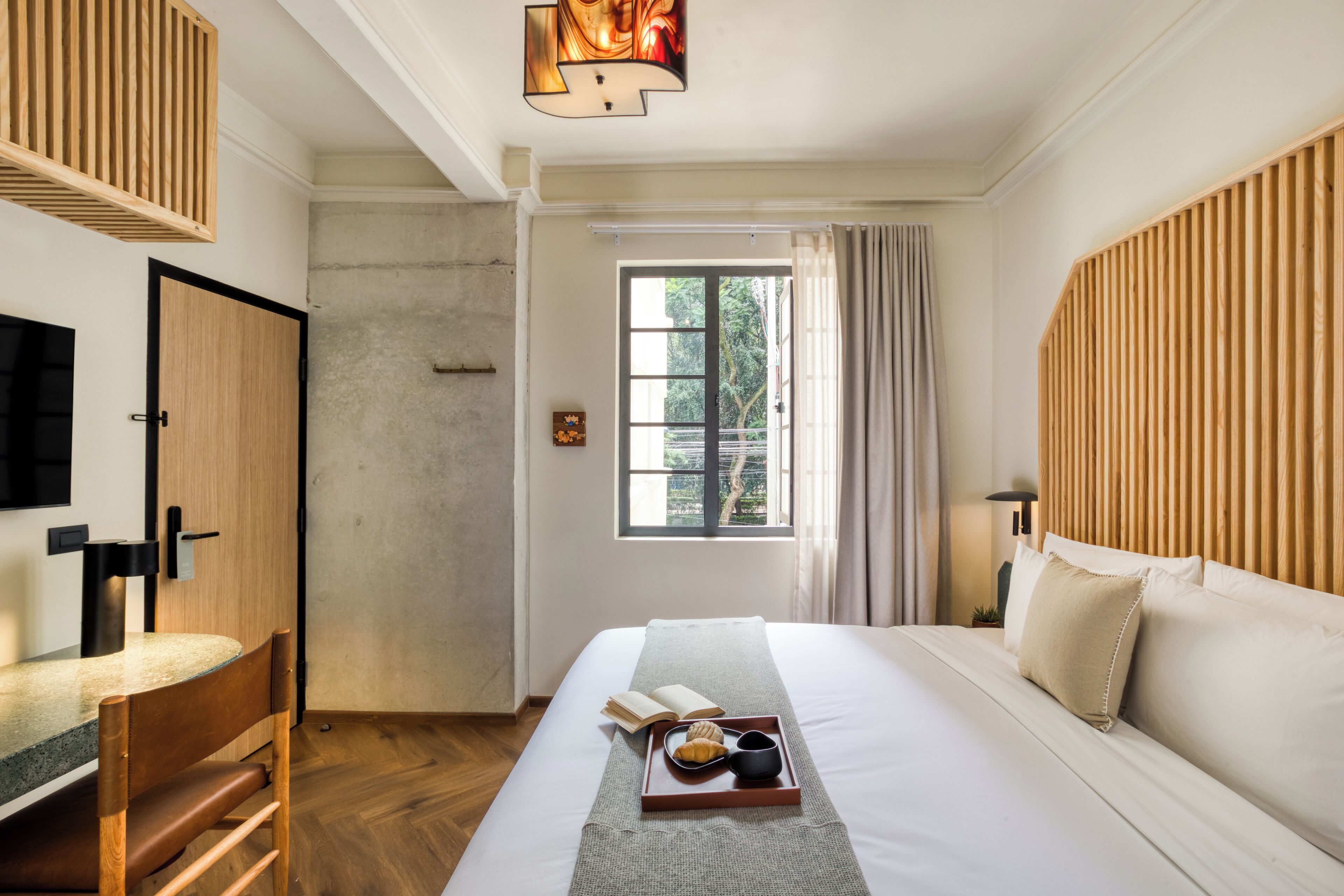 Quarto de hotel com cama, cabeceira de madeira, janela com cortinas, mesa de cabeceira com abajur e bandeja com comida e um livro.