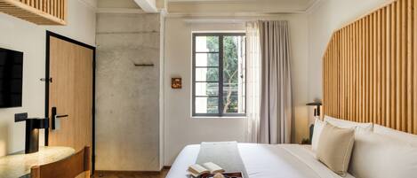Quarto de hotel com cama, cabeceira de madeira, janela com cortinas, mesa de cabeceira com abajur e bandeja com comida e um livro.