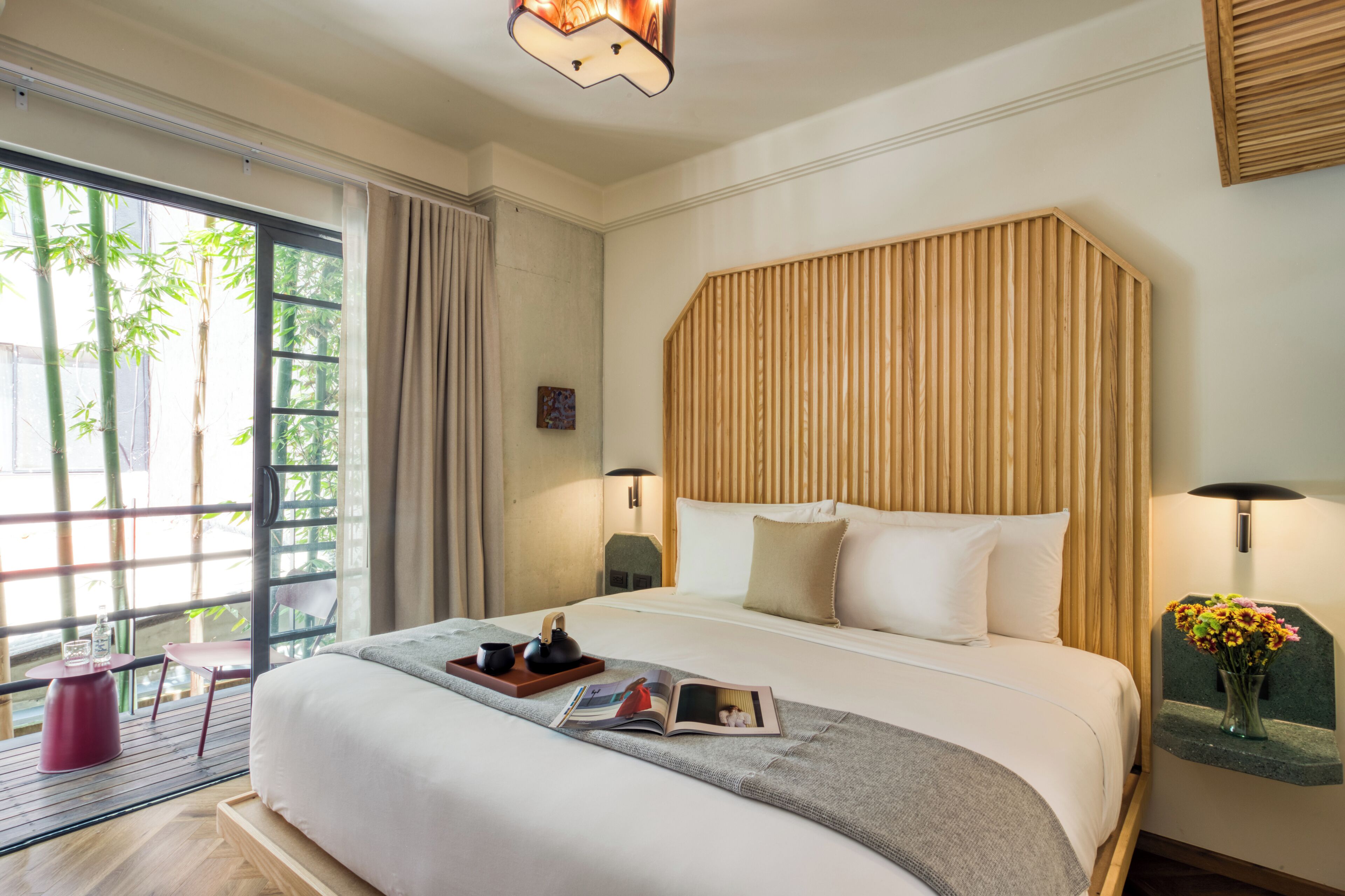 Quarto de hotel com uma cama grande, uma mesinha de cabeceira com abajur, uma pequena mesa com um bule e uma revista, e vista para o verde lá fora.